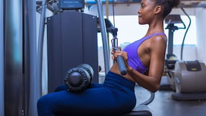 Uma pessoa usando uma máquina de leg press em uma academia, com equipamentos de ginástica ao fundo.