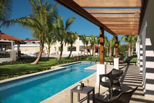Uma área à beira da piscina com um pérgola de madeira, espreguiçadeiras e uma mesa com uma bebida.