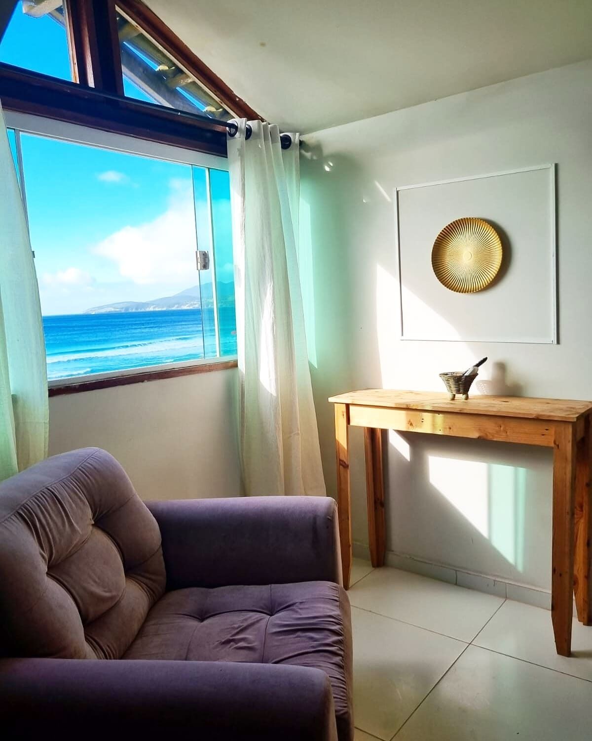 Quarto com vista para o oceano, uma poltrona estofada, uma mesa de madeira e um prato decorativo na parede.