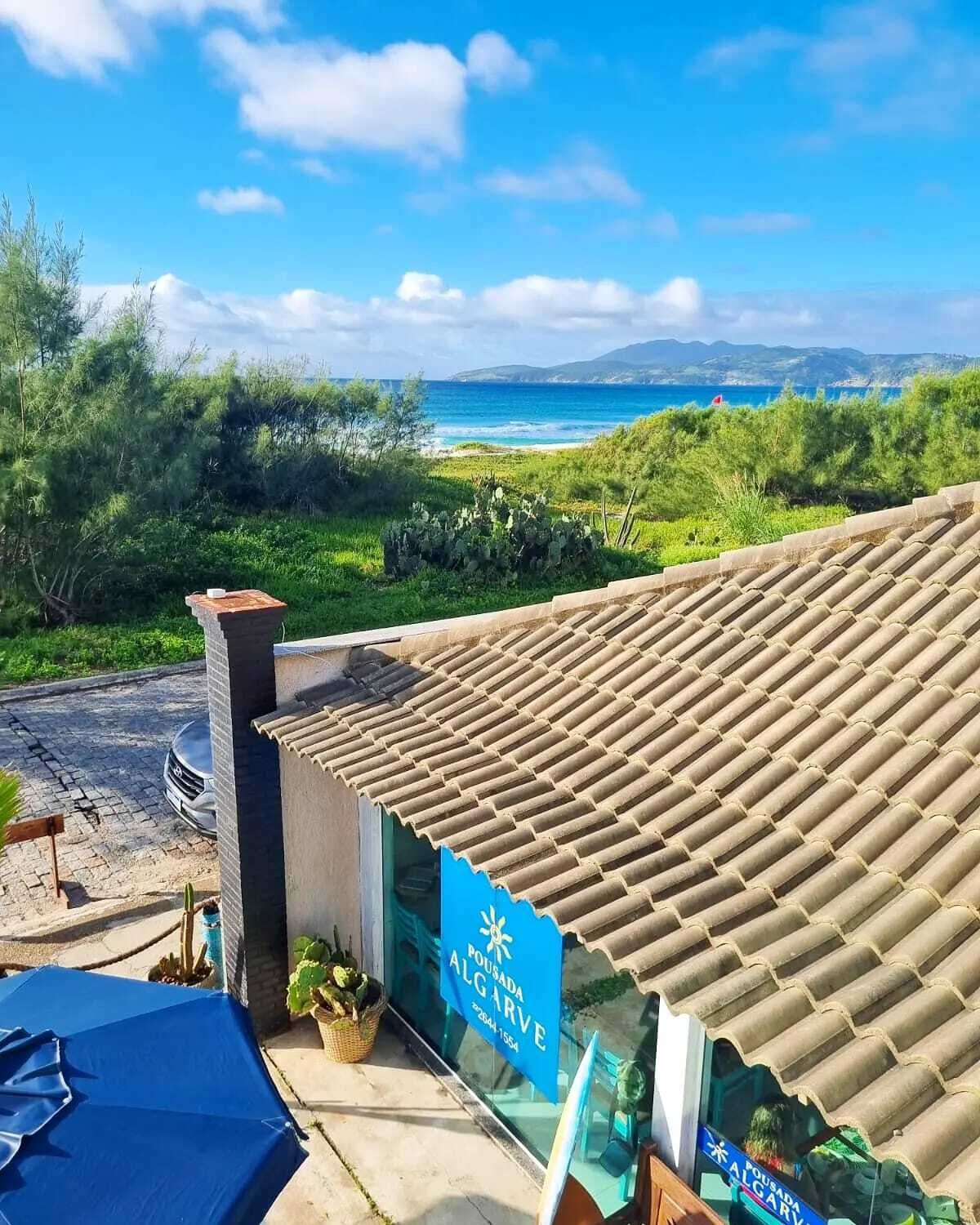 Terraço no telhado com vista para o mar e colinas, um letreiro com 'POUSADA ALGARVE' e um carro estacionado.