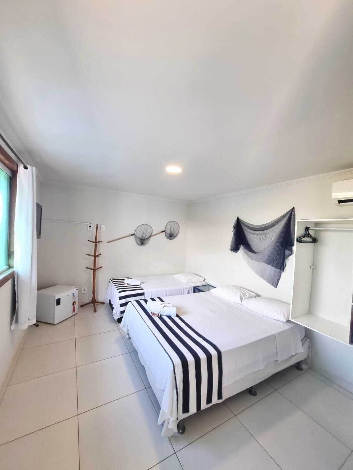 Quarto de hotel com duas camas de solteiro, uma mesinha, uma cadeira, um cabideiro e uma janela com cortinas.