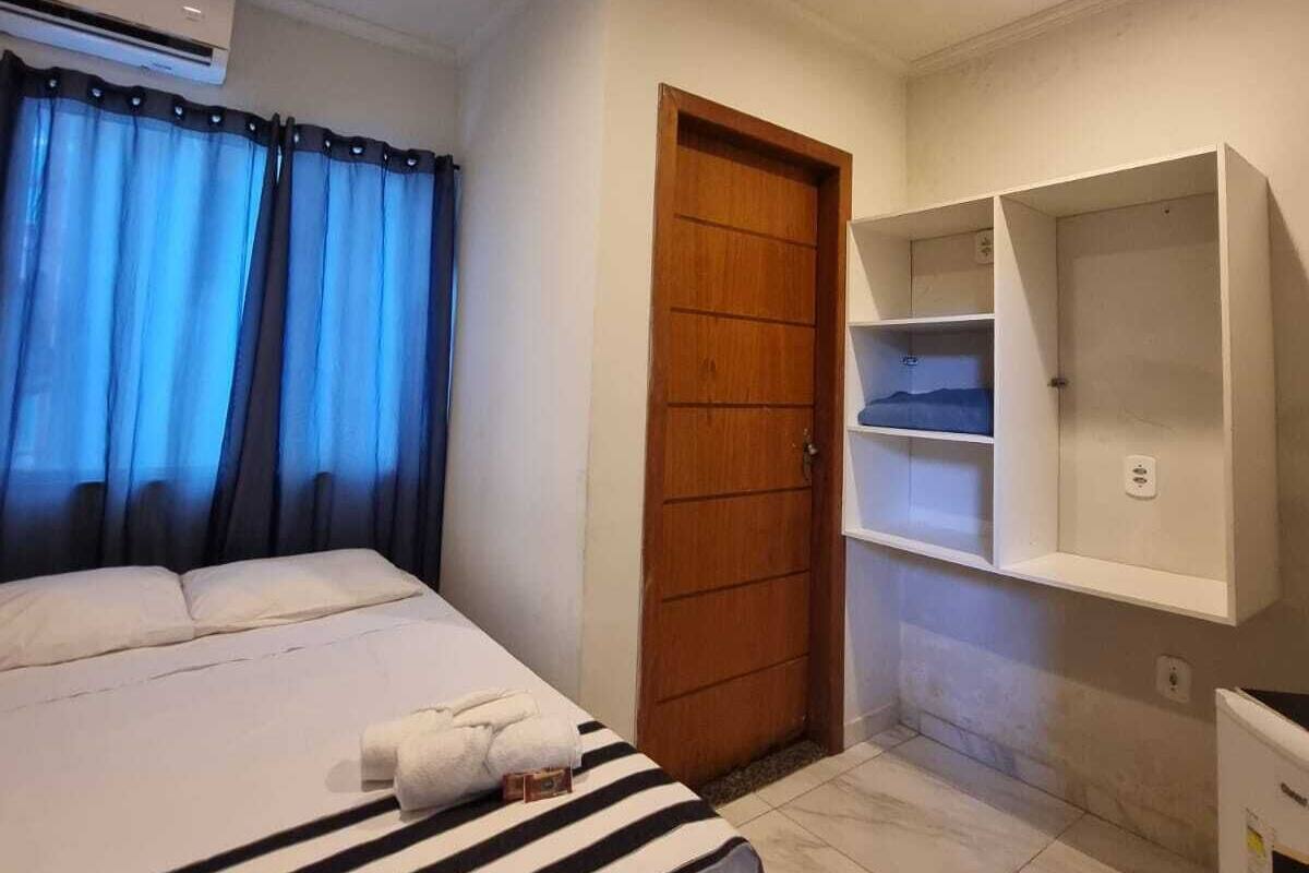 Quarto de hotel com uma cama, porta de madeira, estante branca e unidade de ar condicionado.