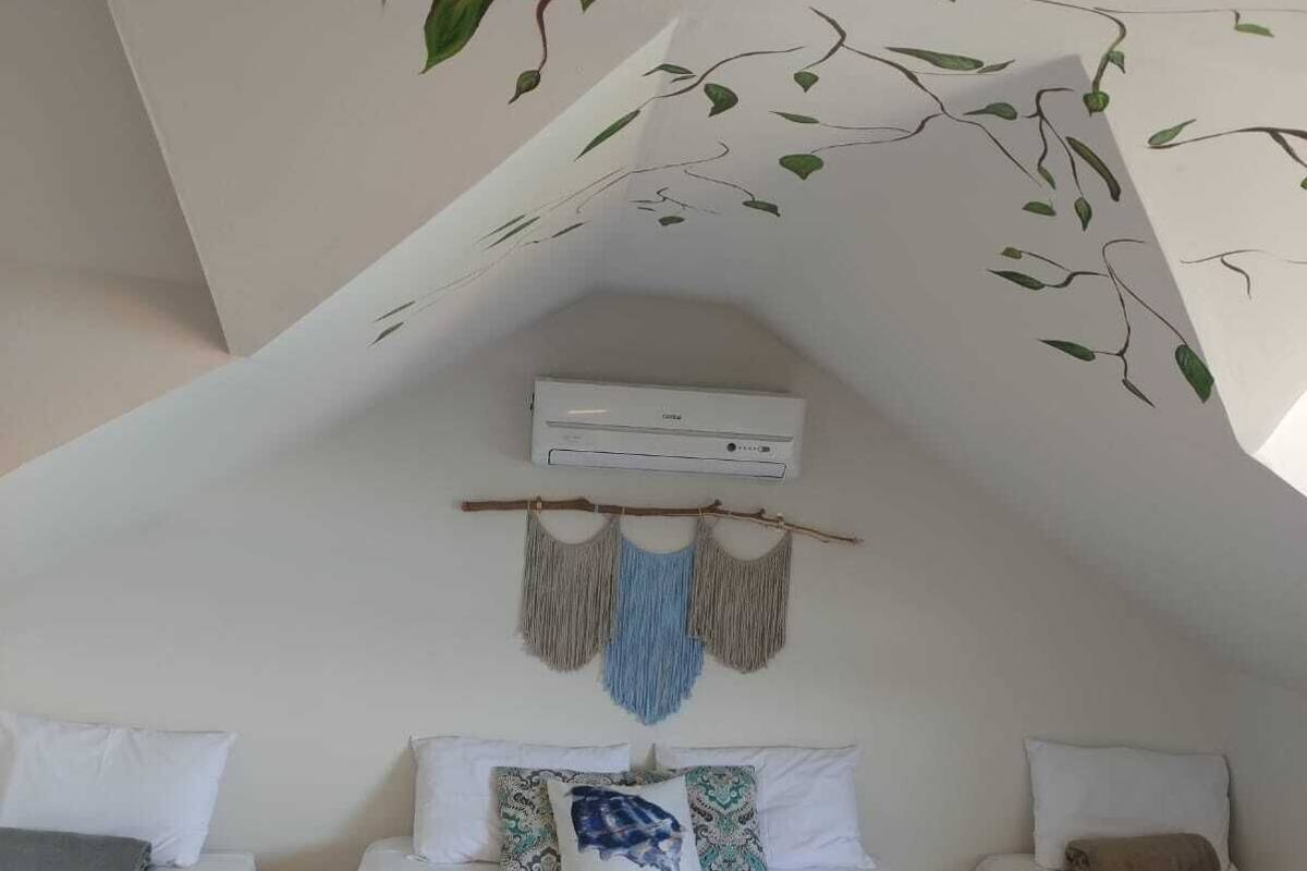 Quarto com teto inclinado, uma cama com colcha verde, duas mesas de cabeceira e um ar condicionado fixado na parede.