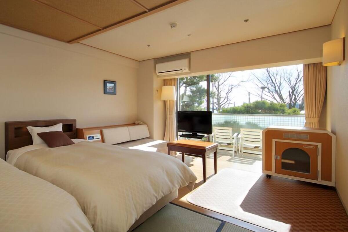 Quarto de hotel com duas camas, televisão, uma mesinha e vista para o exterior.