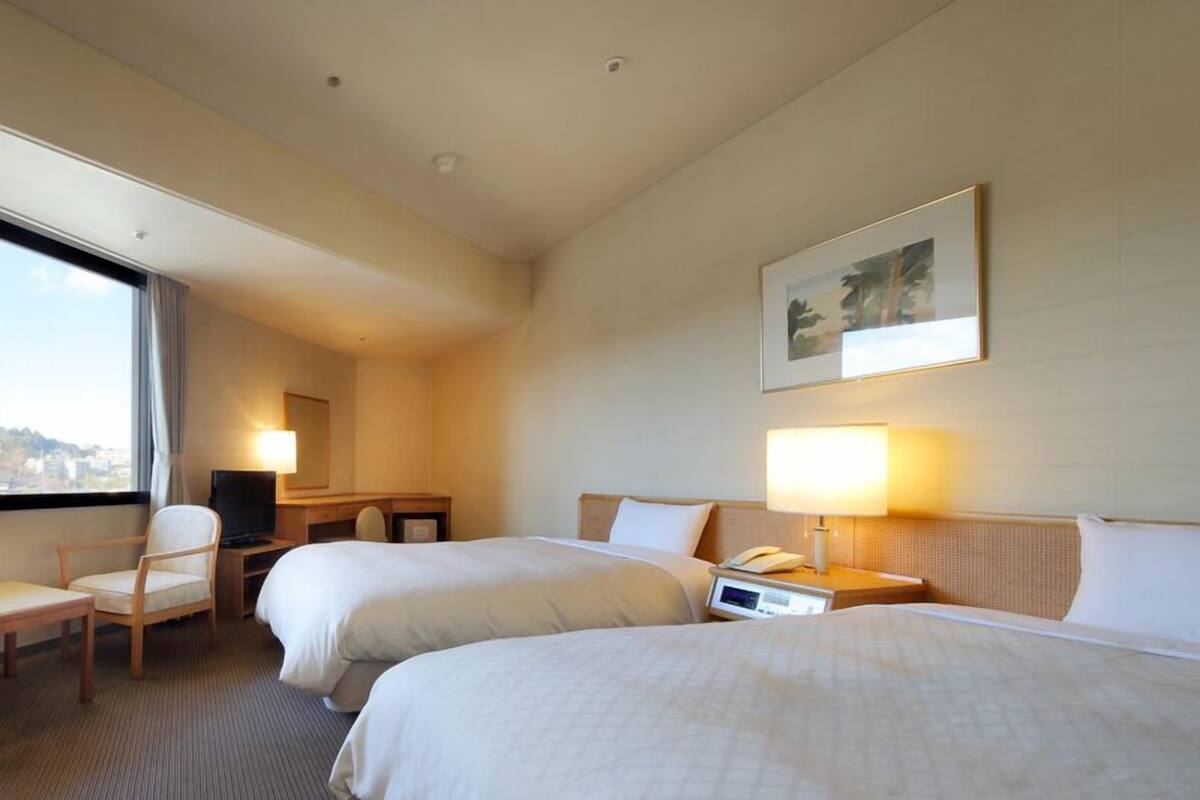 Quarto de hotel com duas camas, uma escrivaninha, uma cadeira, televisão e janela com vista.