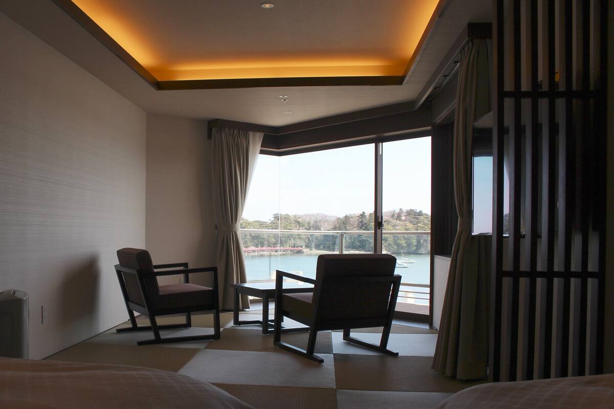 Quarto de hotel com cama, duas cadeiras, mesa e uma janela grande com vista para a água.