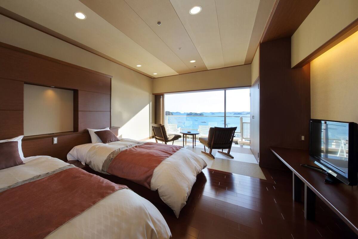 Quarto de hotel com duas camas, televisão, varanda com vista para o mar e área de refeições.