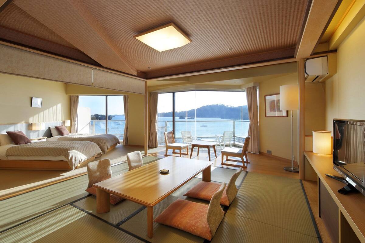 Quarto de hotel com cama, mesa de jantar, cadeiras, televisão e vista para o mar.