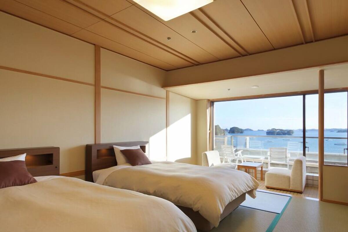Quarto de hotel com duas camas, uma janela grande com vista para o mar e um pequeno espaço de estar.