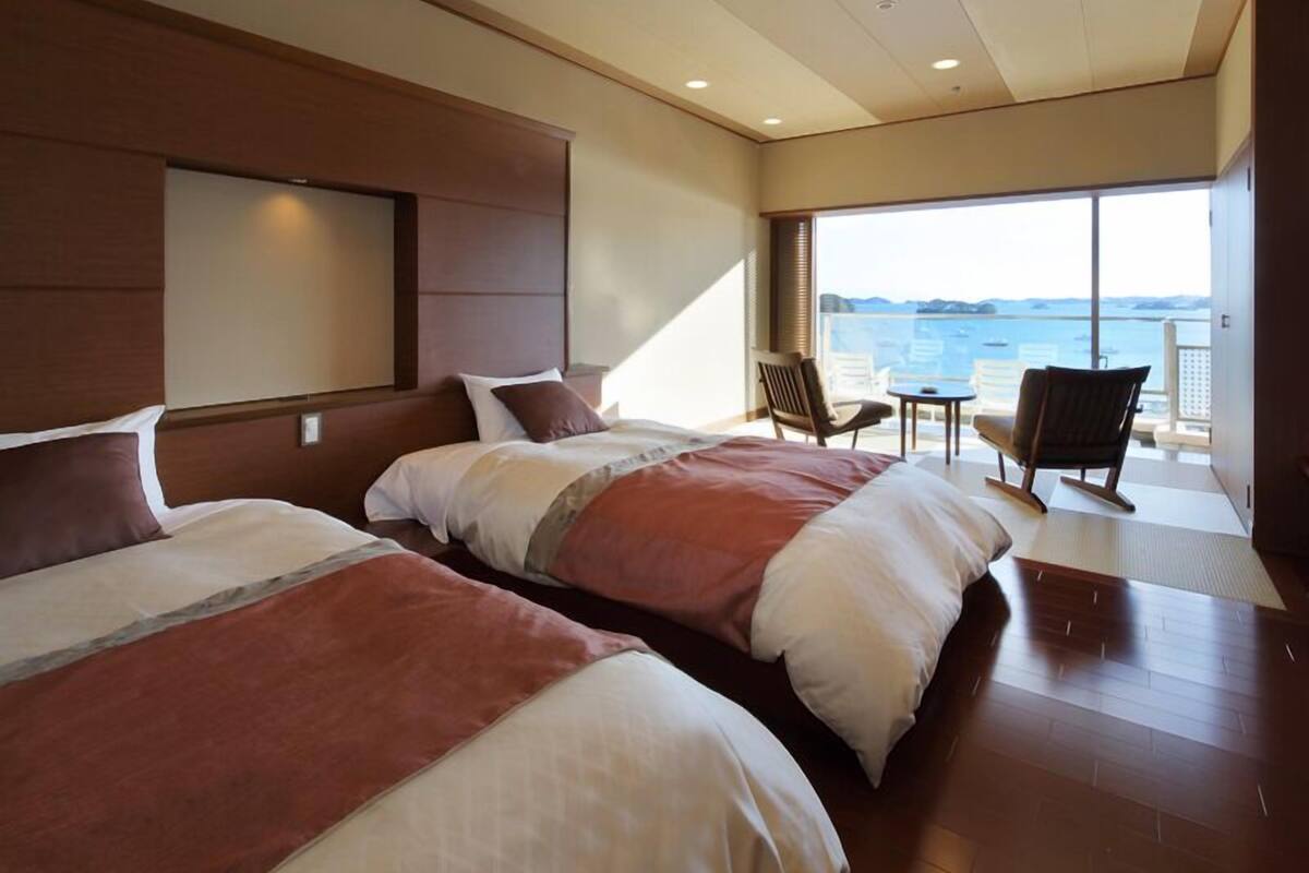 Quarto de hotel moderno com duas camas, uma janela grande com vista para o mar e uma varanda com assentos.