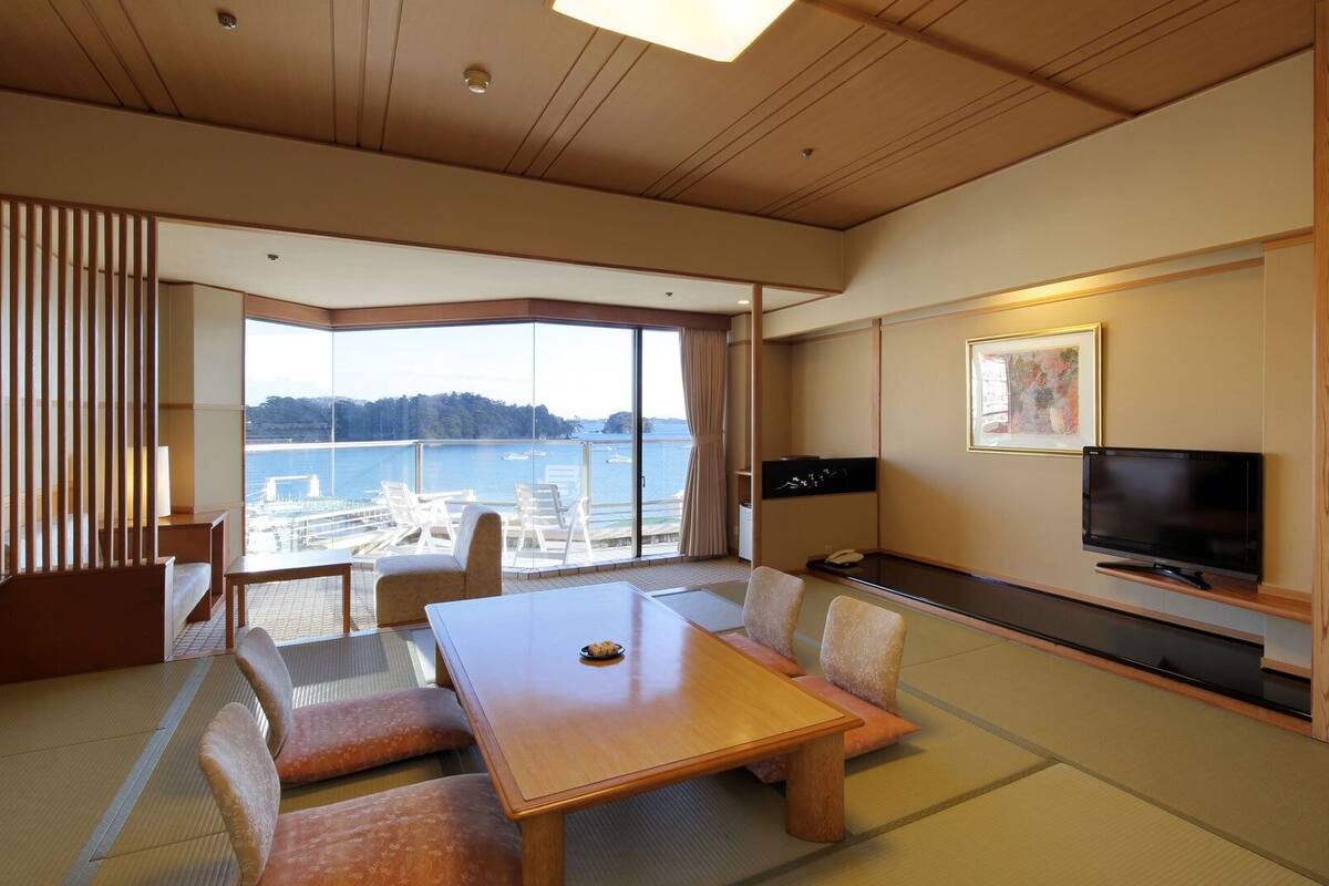 Quarto de hotel com piso de tatame, uma mesa baixa, assentos acolchoados, televisão e uma grande janela com vista para o mar.