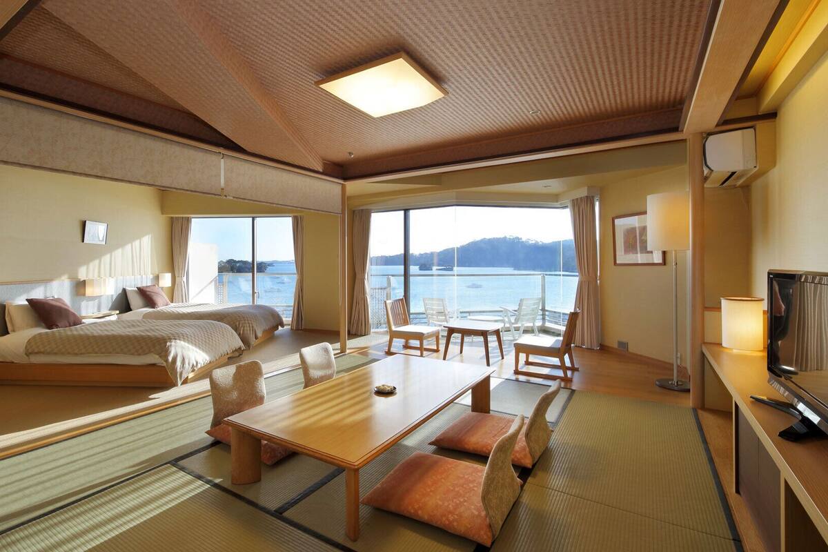 Quarto de hotel com cama, mesa de jantar, cadeiras, televisão e vista para o mar.