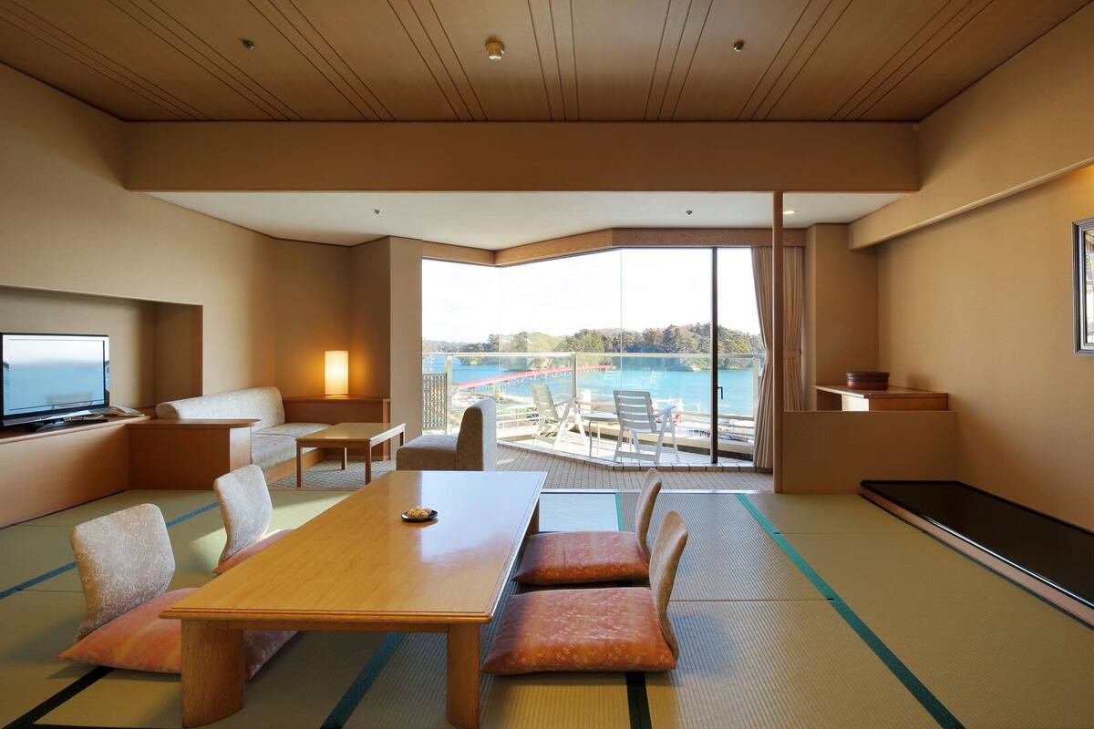 Quarto de hotel com piso de tatame, mesa de jantar, cadeiras, televisão, sofá, varanda com vista para o mar e uma grande janela.
