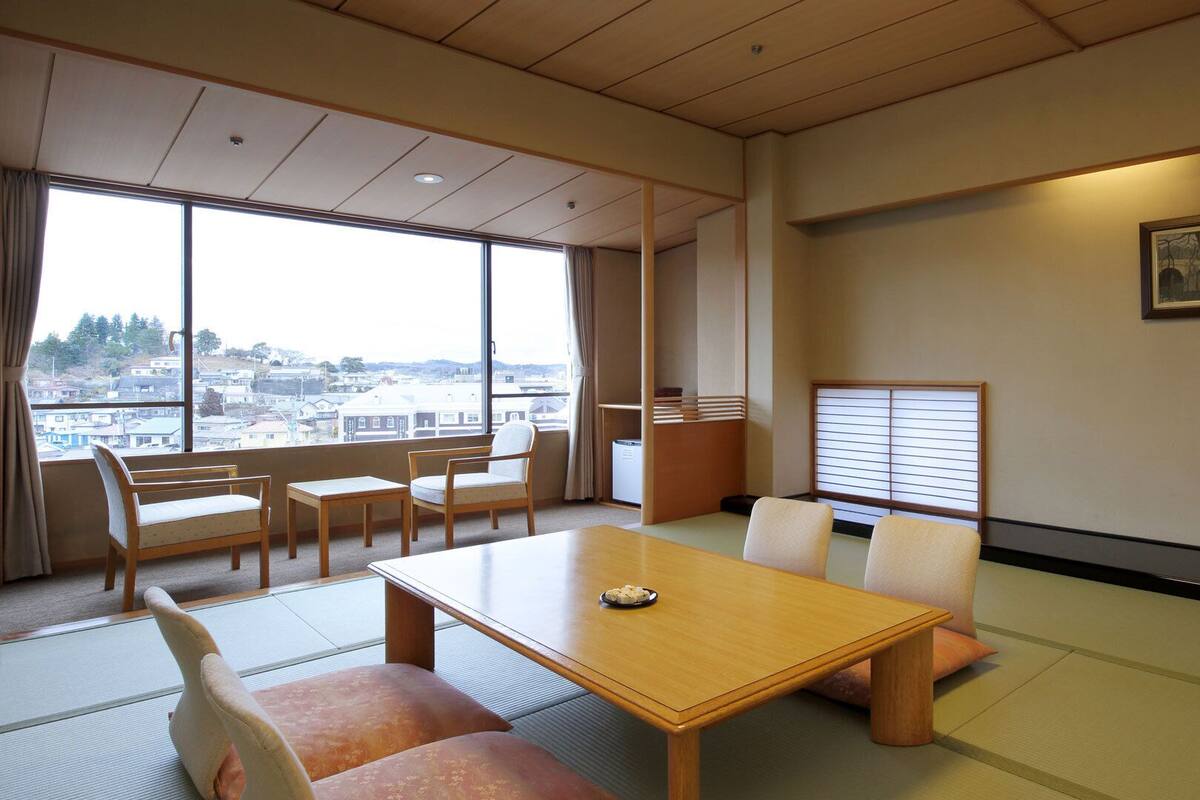 Quarto de hotel com tatames, uma mesa de madeira, cadeiras, vista da cidade e um quadro na parede.