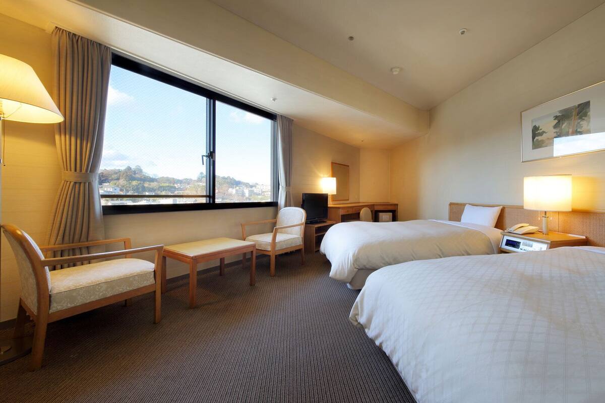 Quarto de hotel com duas camas, uma escrivaninha, uma cadeira, uma televisão e uma janela com vista.