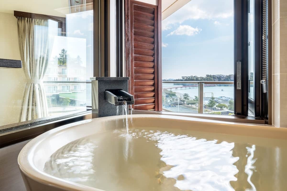 Um banheiro com banheira, janela com vista para uma ponte e água, e um corrimão.