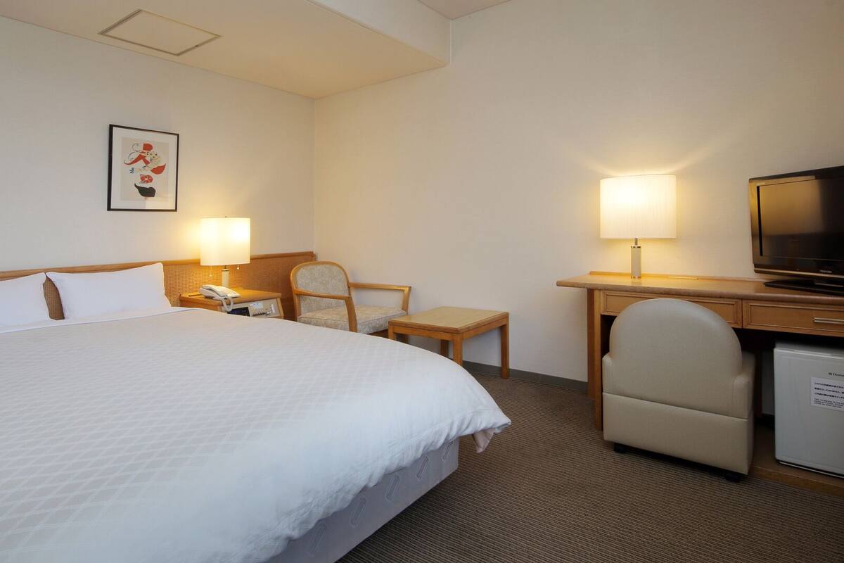 Quarto de hotel com uma cama grande, uma escrivaninha com abajur, uma cadeira, televisão e um pequeno frigorífico.