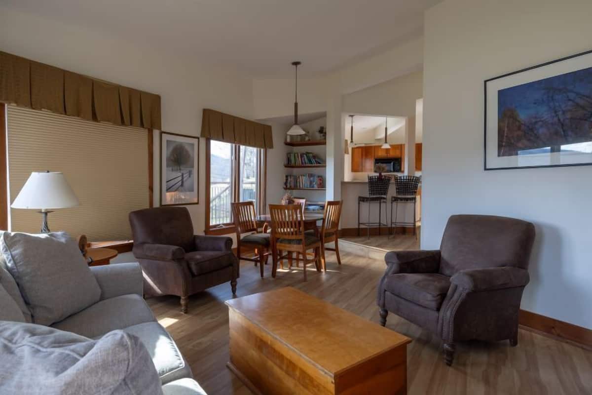 Uma sala de estar com sofá, poltronas, mesa de centro de madeira e um espaço de jantar com cadeiras e mesa. Há uma estante com livros e um quadro na parede.