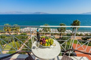 Uma varanda com uma mesa posta, com frutas e copos de vinho, com vista para a praia e o mar.