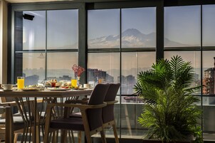 Uma área de refeições com mesa posta para o café da manhã, grandes janelas com vista para as montanhas e um vaso de planta.