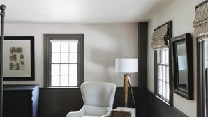 Quarto com piso de madeira, uma poltrona branca, um abajur e um quadro na parede.