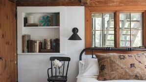 Quarto com teto de madeira, uma cama, uma cadeira, uma estante com livros e uma janela.
