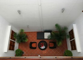 Área externa coberta com parede de azulejos, duas plantas em vasos, um objeto circular preto e uma parede branca com uma janela.
