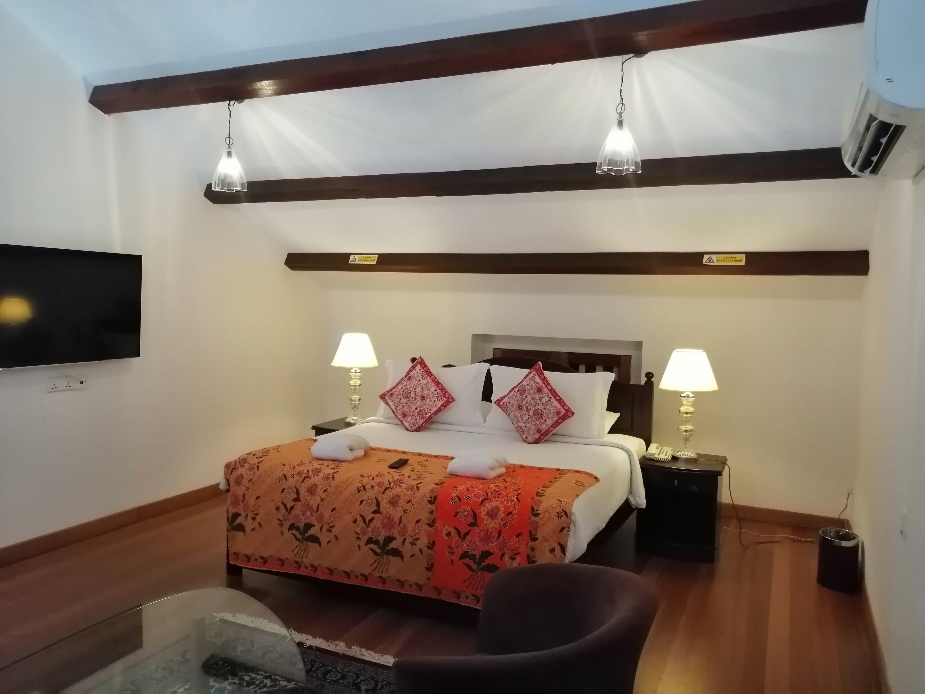 Quarto de hotel com cama, duas luminárias laterais, televisão presa na parede e ventilador de teto.