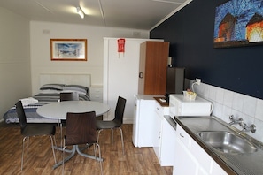 Quarto compacto com uma cama, uma pequena mesa de jantar com cadeiras, um espaço de cozinha com pia e uma geladeira.