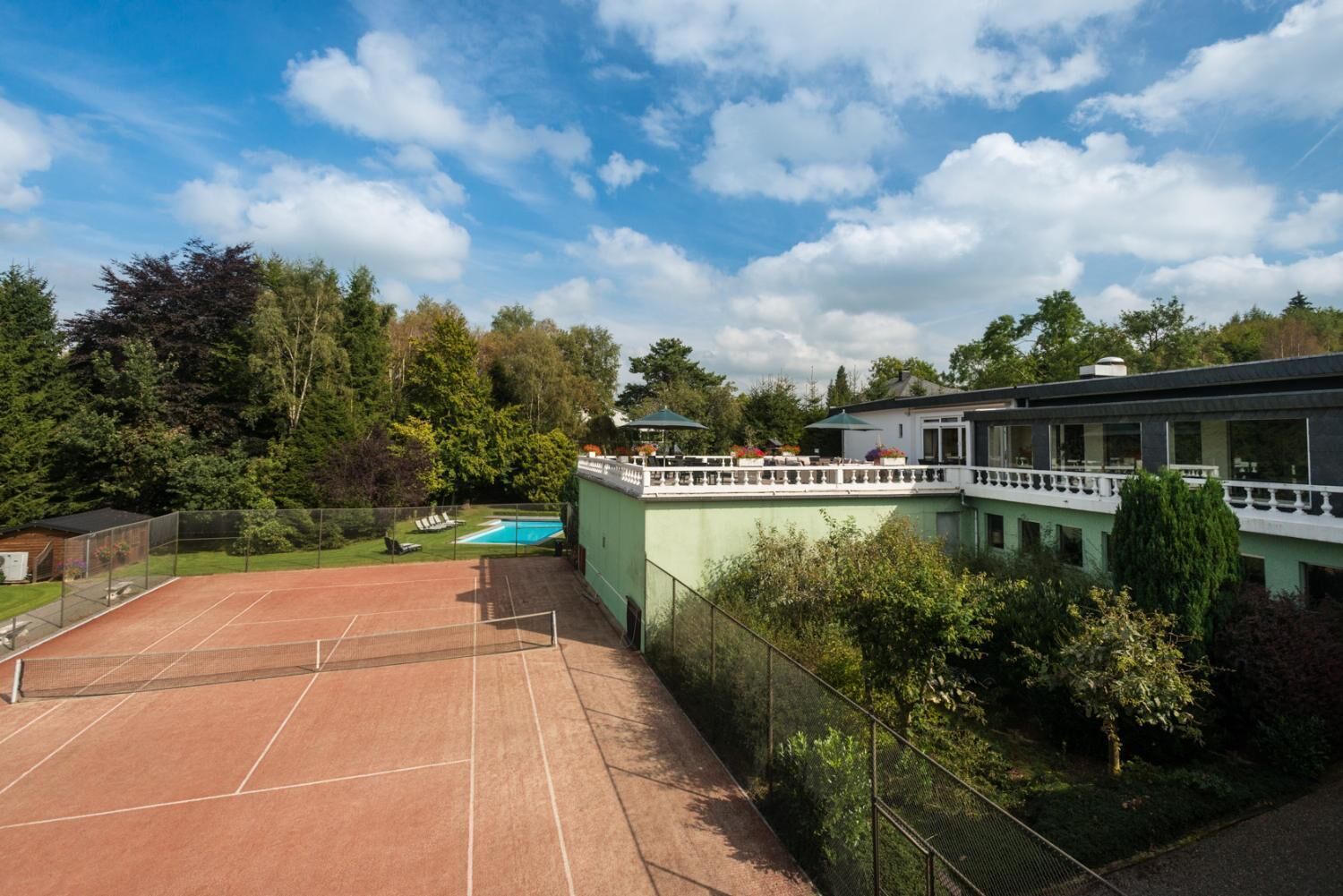 Uma quadra de tênis com piscina, rodeada por vegetação e um edifício com varanda.