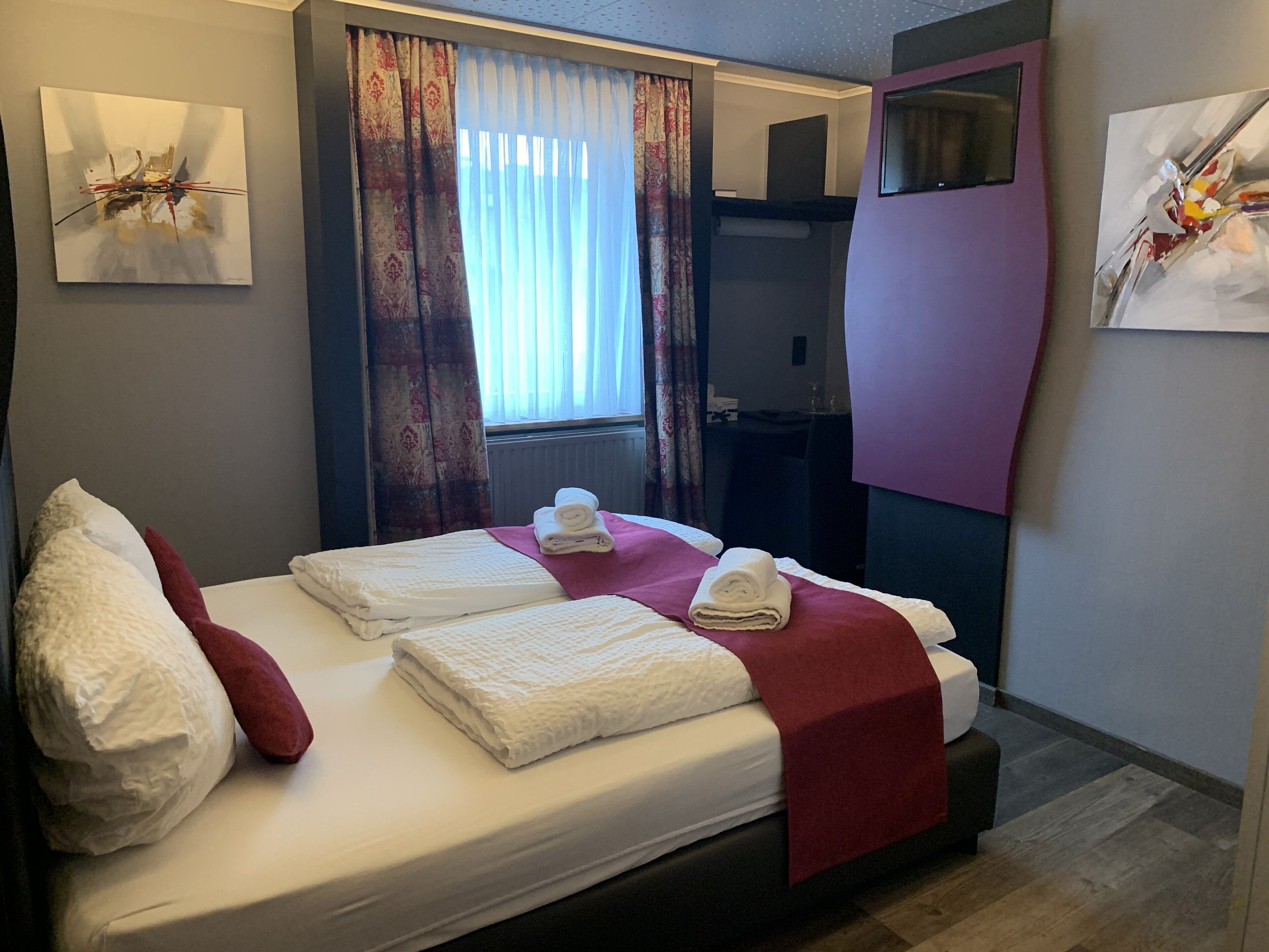 Uma cama bem arrumada com lençóis brancos e detalhes vermelhos, uma janela com cortinas leves e um espelho na parede.