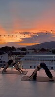 Duas pessoas praticando yoga em tapetes ao ar livre, com paisagem urbana e montanhas ao fundo.