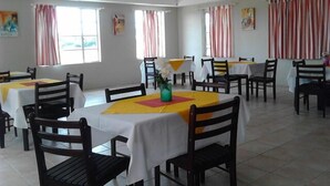 Uma sala de jantar com várias mesas e cadeiras, janelas amplas com cortinas e um candelabro.