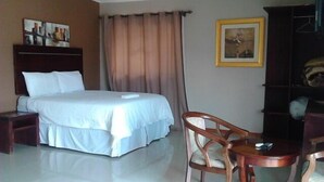 Quarto de hotel com cama, uma cadeira de madeira, mesinha, mesa de cabeceira, guarda-roupa e um quadro na parede.