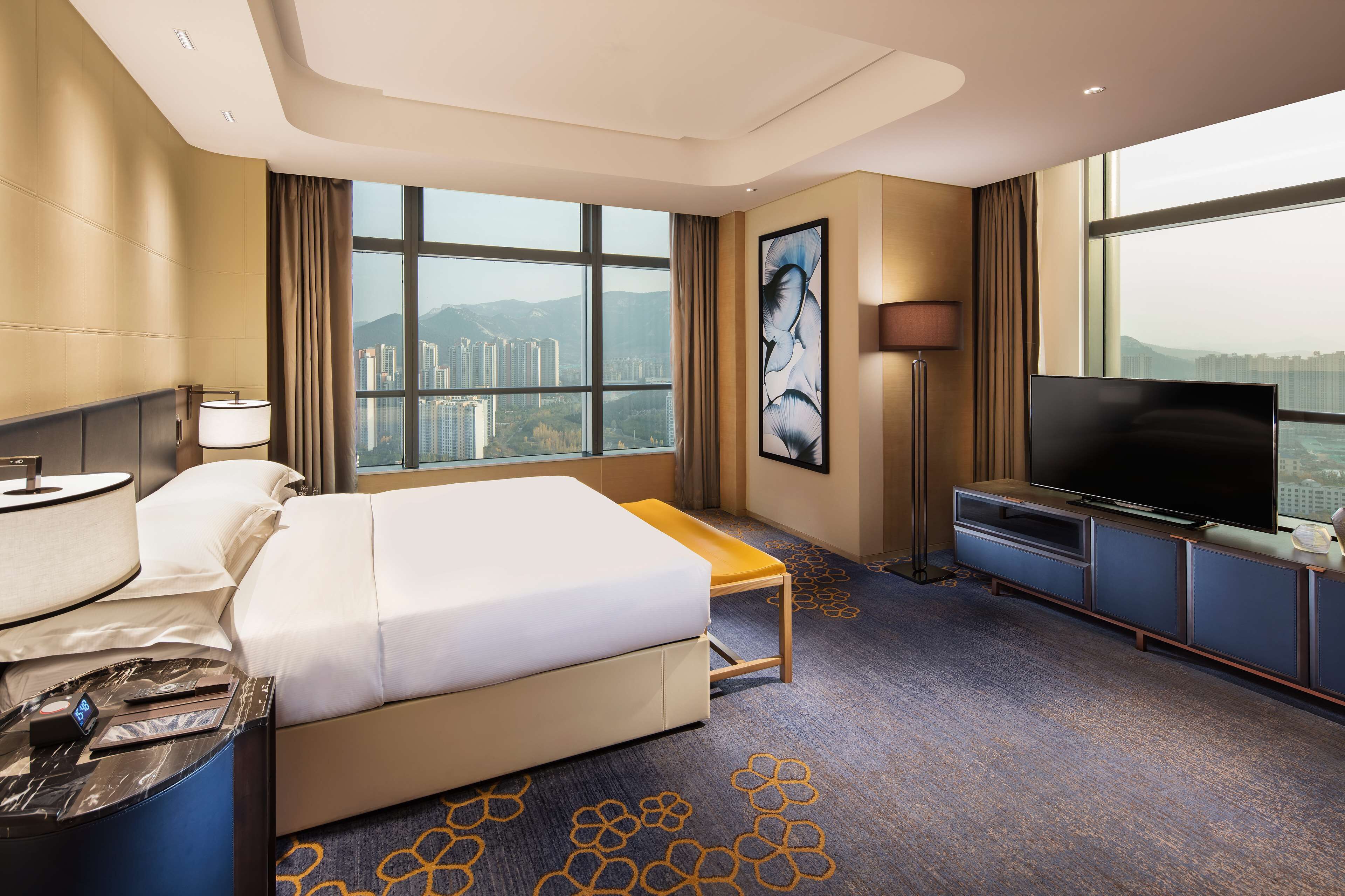Quarto de hotel com uma cama grande, televisão, secretária e vista para a paisagem urbana através de janelas amplas.