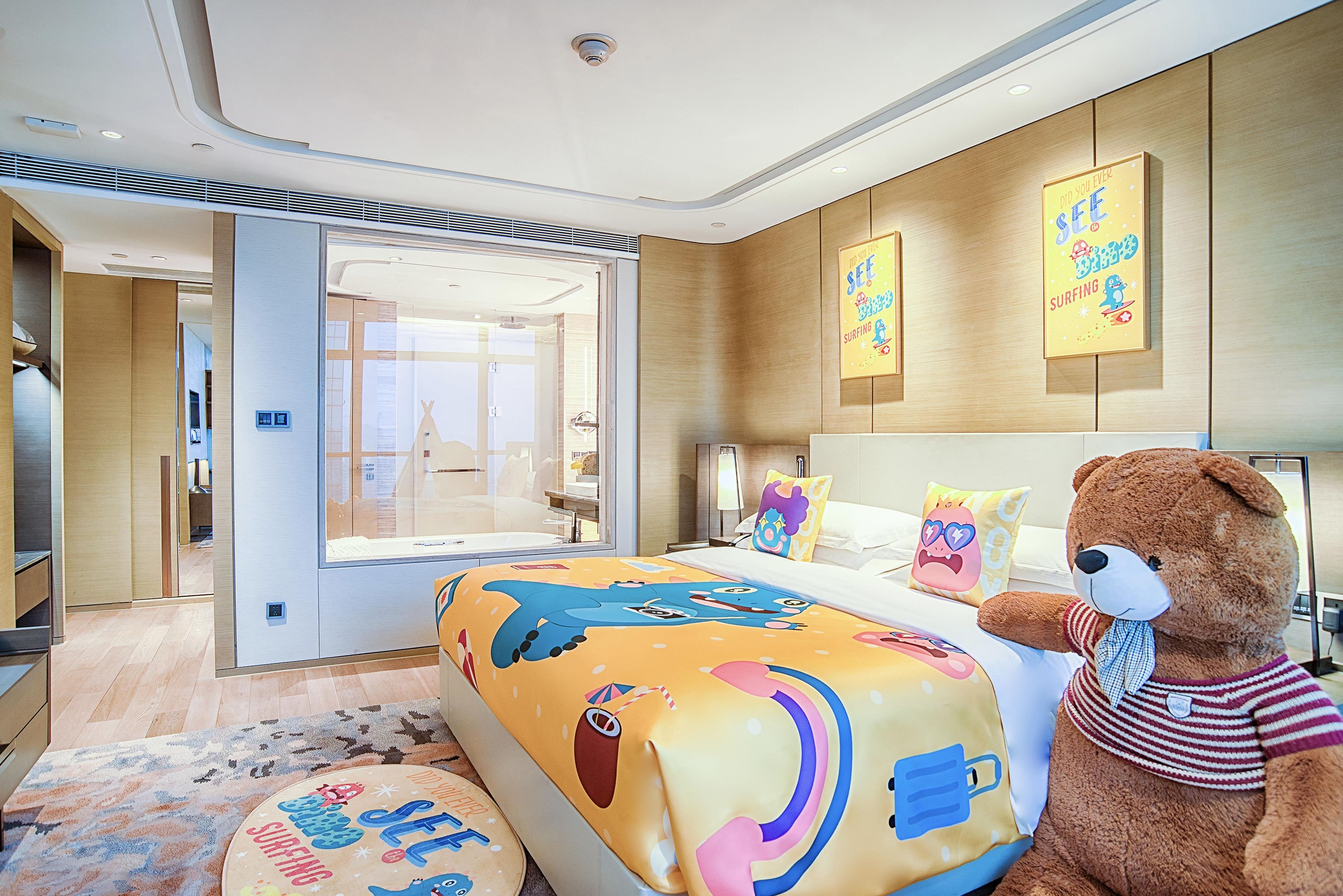 Quarto de hotel com cama, um urso de pelúcia, uma janela grande e um quadro na parede.