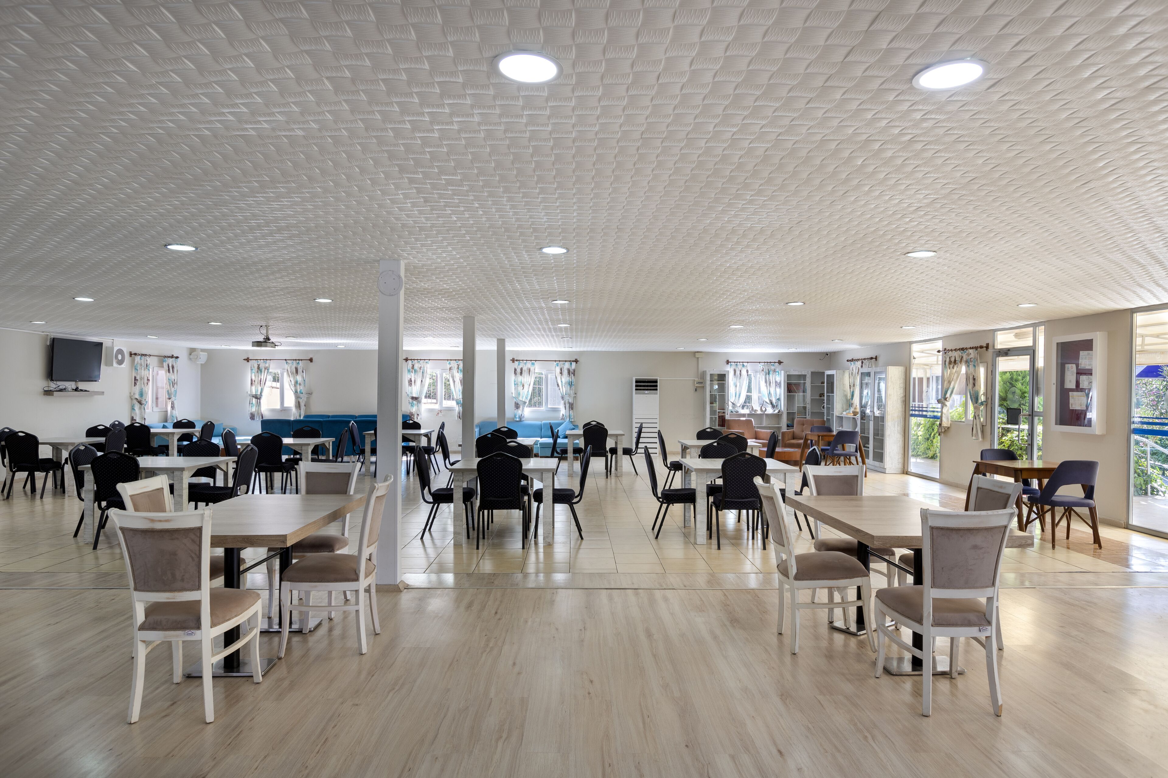 Uma sala de jantar ampla, com várias mesas e cadeiras, teto estampado e grandes janelas que proporcionam iluminação natural.