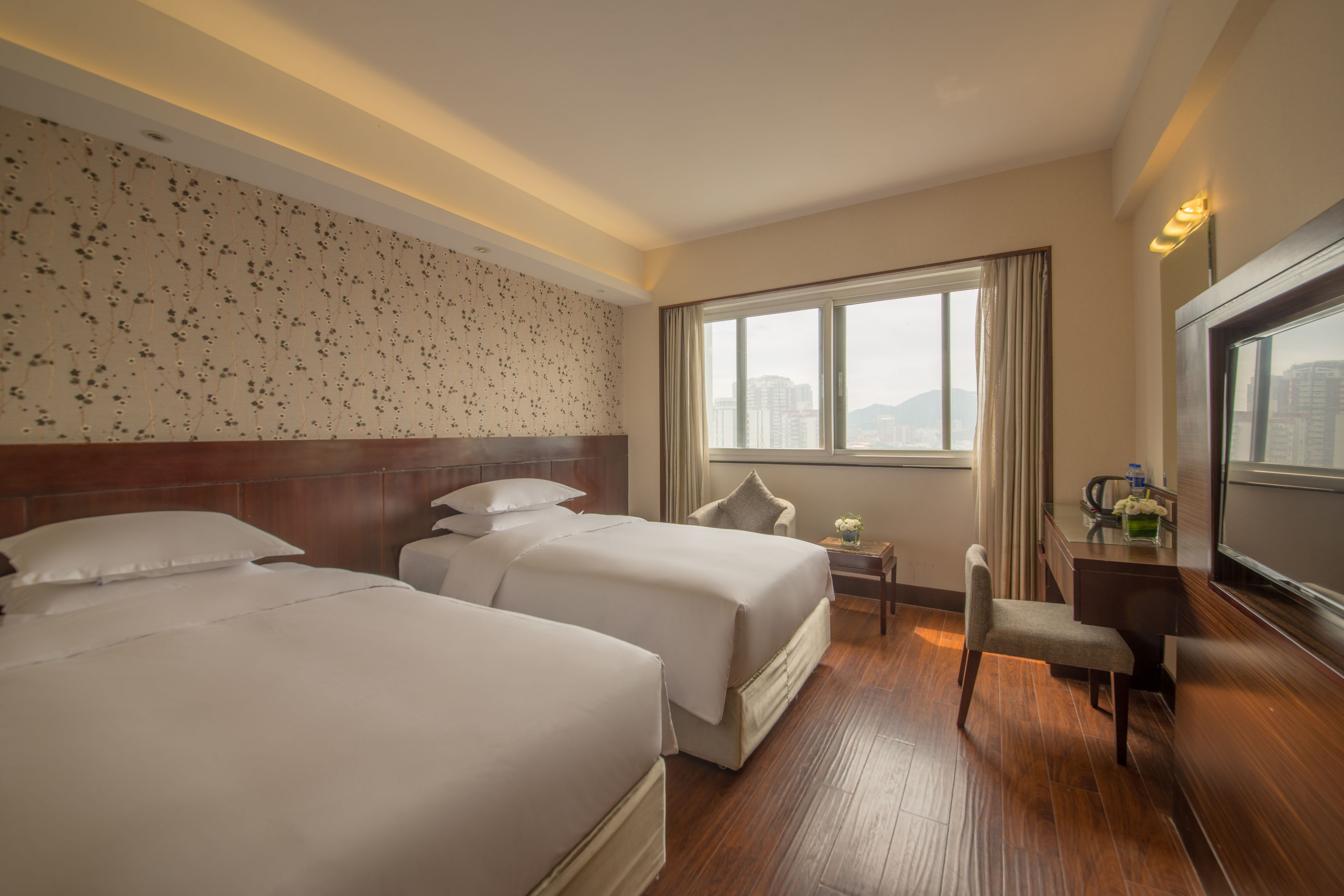 Quarto de hotel com duas camas, uma escrivaninha, uma cadeira, janela com vista para a cidade e parede decorativa.