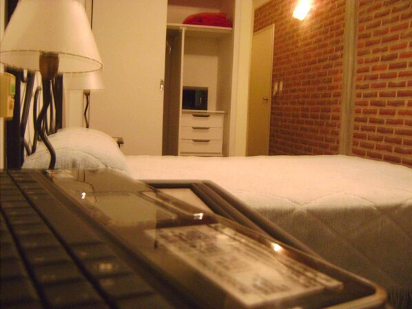 Quarto de hotel com uma cama, uma escrivaninha com um laptop, uma luminária, um guarda-roupa e uma parede de tijolos.