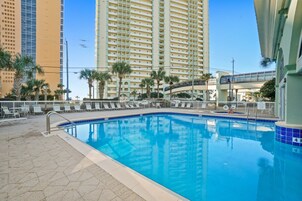 Piscina com espreguiçadeiras e palmeiras, cercada por edifícios altos e uma ponte para pedestres.