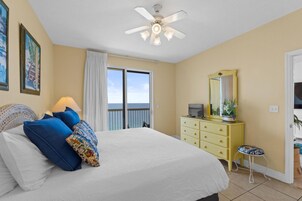 Quarto com cama, cômoda, espelho, ventilador de teto e vista para o oceano.
