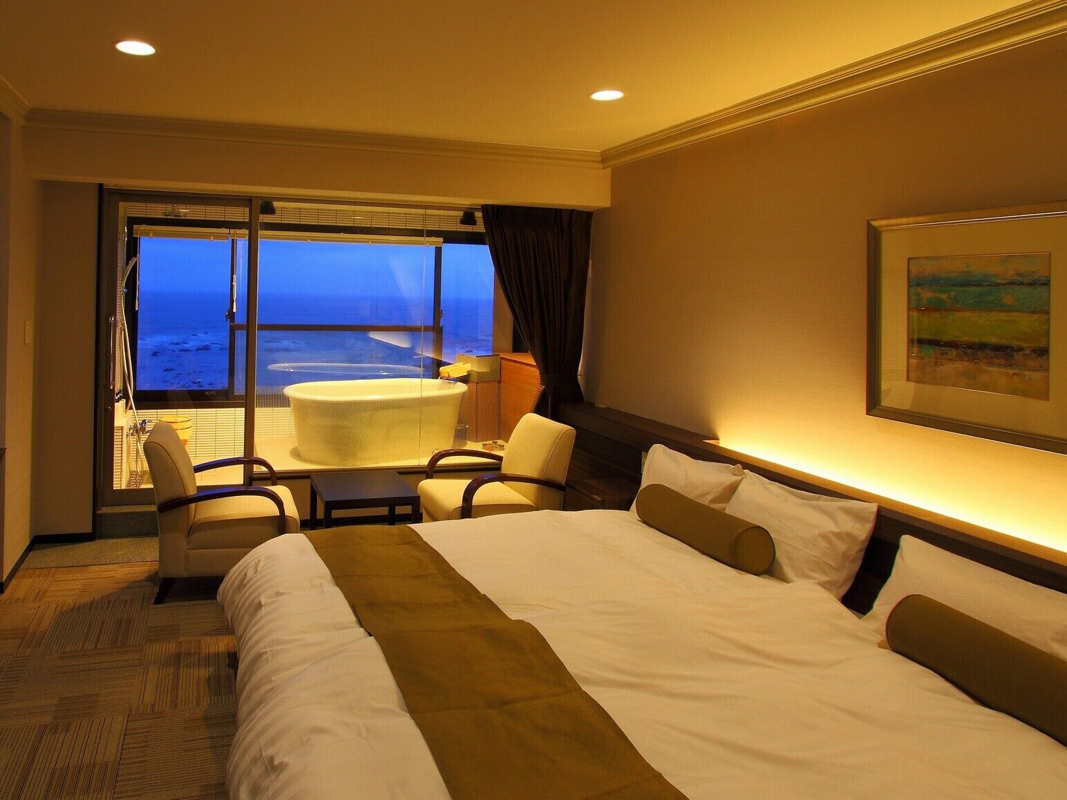 Quarto de hotel com uma cama grande, área de estar com cadeiras, uma mesinha, banheira e uma janela ampla com vista para o oceano.