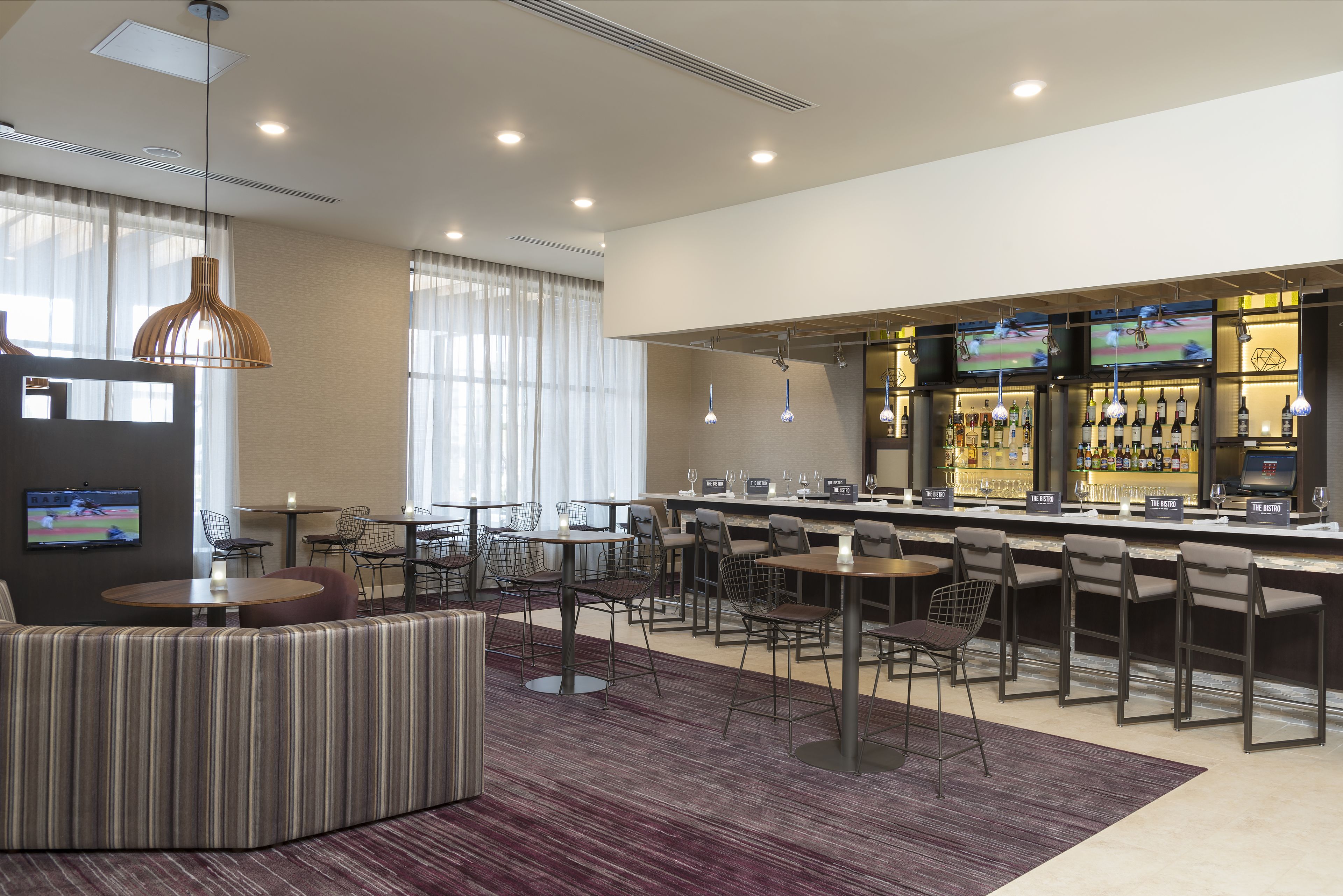 Uma área de bar moderna com diversas opções de assento, incluindo banquetas e mesas, e uma tela de televisão fixada na parede.