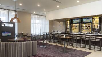 Uma área de bar moderna com diversas opções de assento, incluindo banquetas e mesas, e uma tela de televisão fixada na parede.