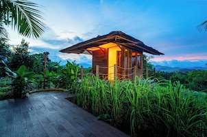 Uma cabana de madeira com telhado de palha, rodeada por vegetação exuberante, acessível por meio de um deck de madeira.