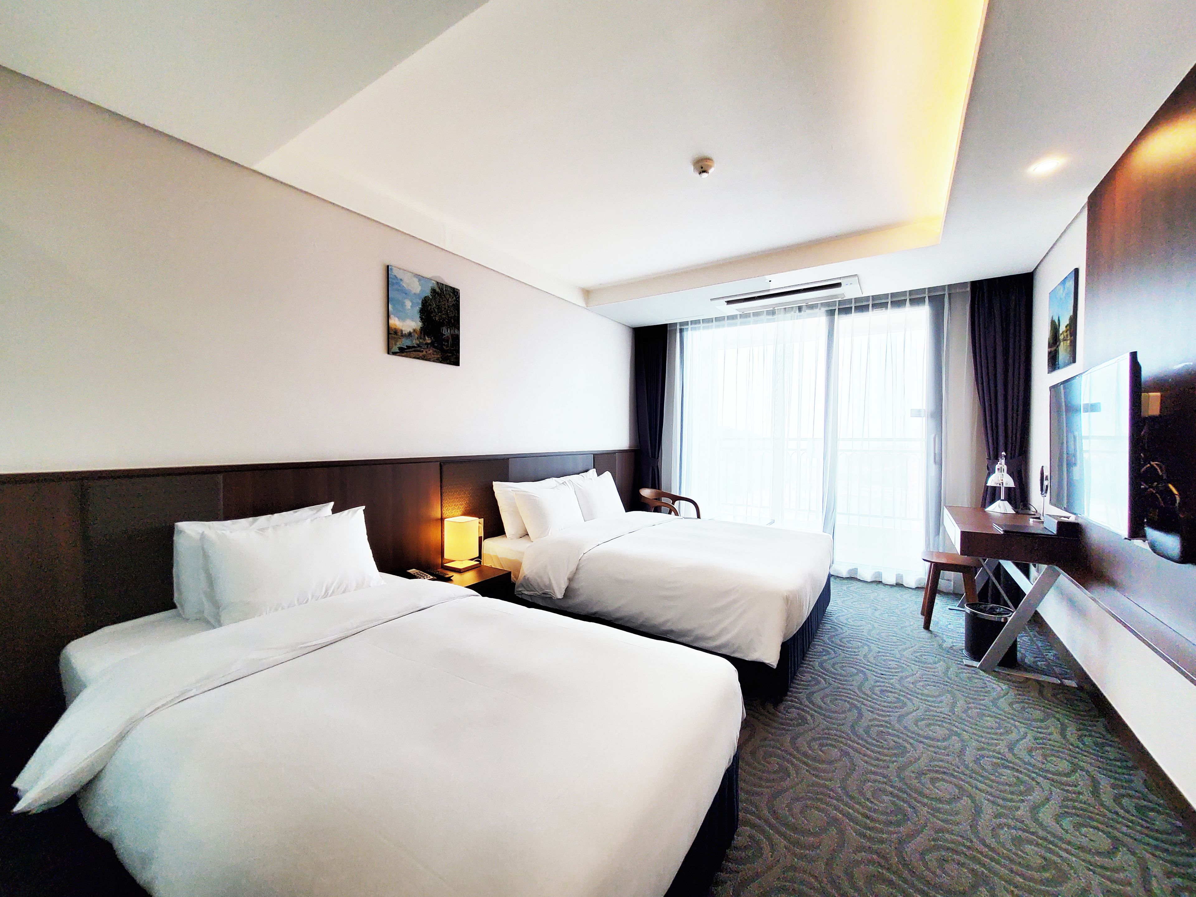 Quarto de hotel com duas camas, uma escrivaninha, uma cadeira, uma televisão e uma janela com cortinas.