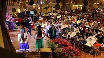 Um evento interno lotado com mesas e cadeiras dispostas em fileiras, um palco com artistas e uma grande plateia.