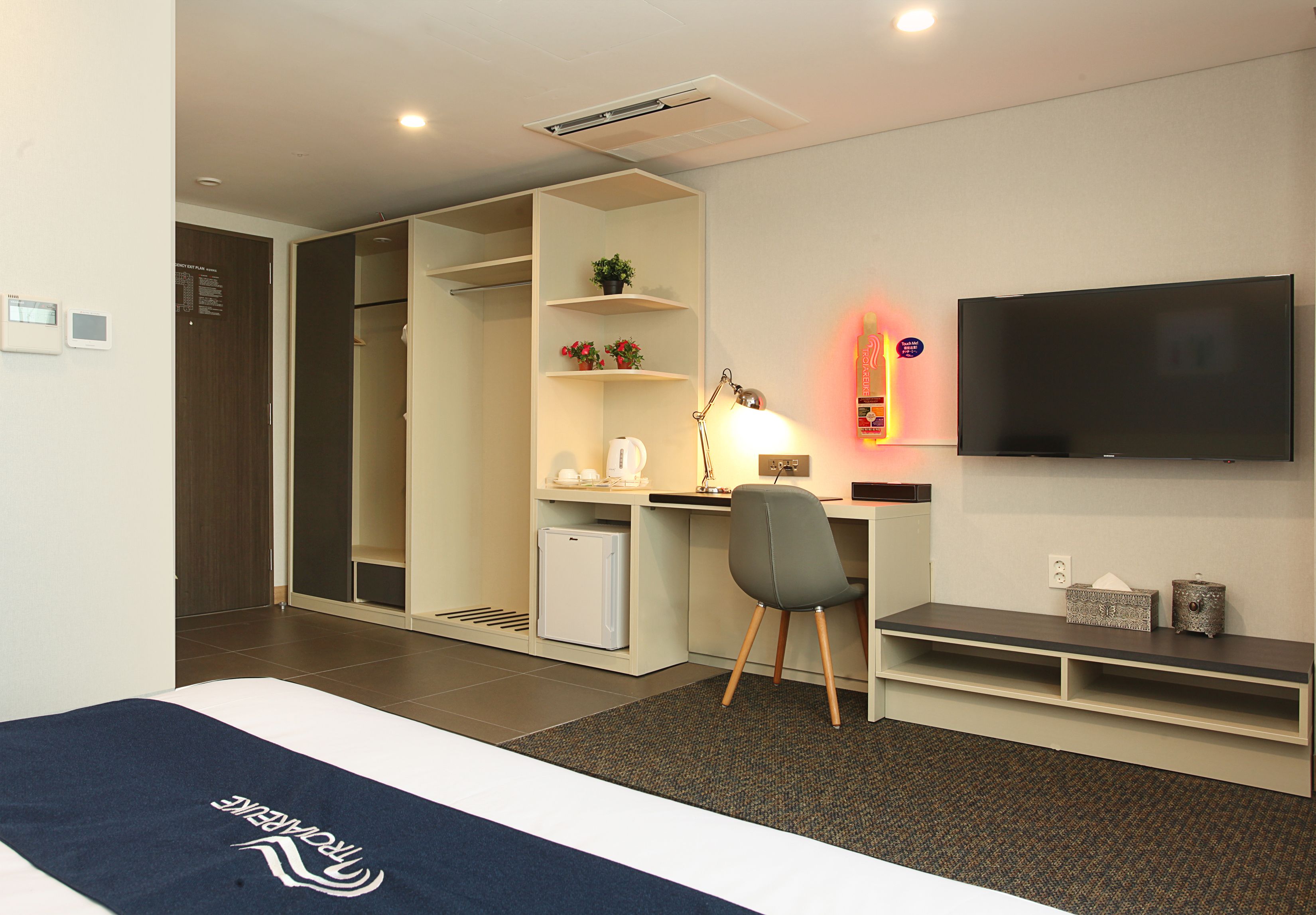 Quarto de hotel com cama, escrivaninha, cadeira, televisão, guarda-roupa e um pequeno frigorífico.