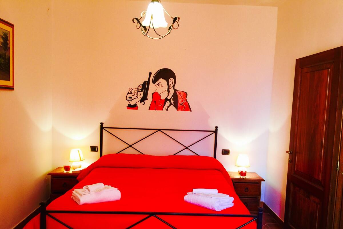 Quarto de hotel com colcha vermelha, cabeceira de metal preta, duas luminárias de apoio e um adesivo de parede com a imagem de um homem e uma mulher.