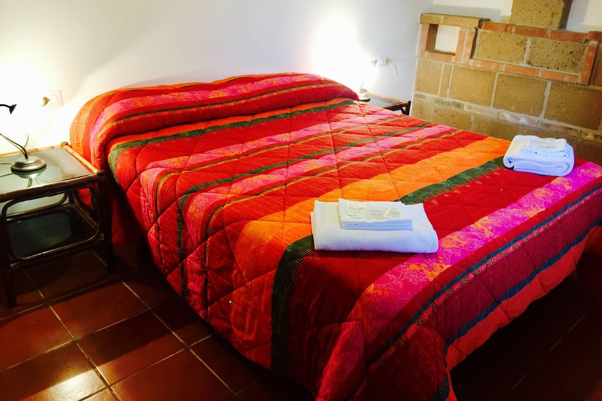 Uma cama com um edredom colorido, uma mesinha de cabeceira com abajur e uma parede de tijolos.